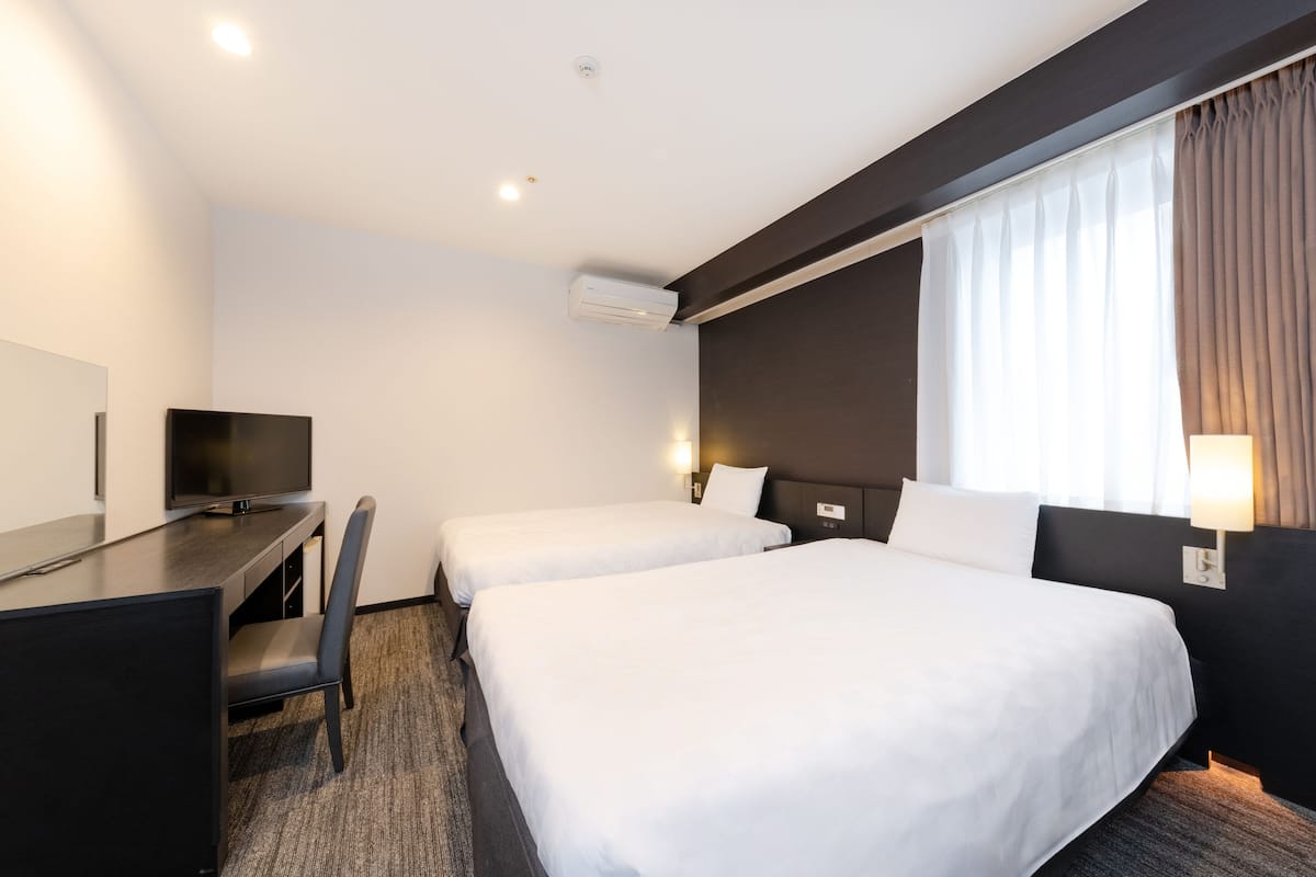 Quarto de hotel com duas camas de solteiro, uma escrivaninha com cadeira, televisão e janela com cortinas.