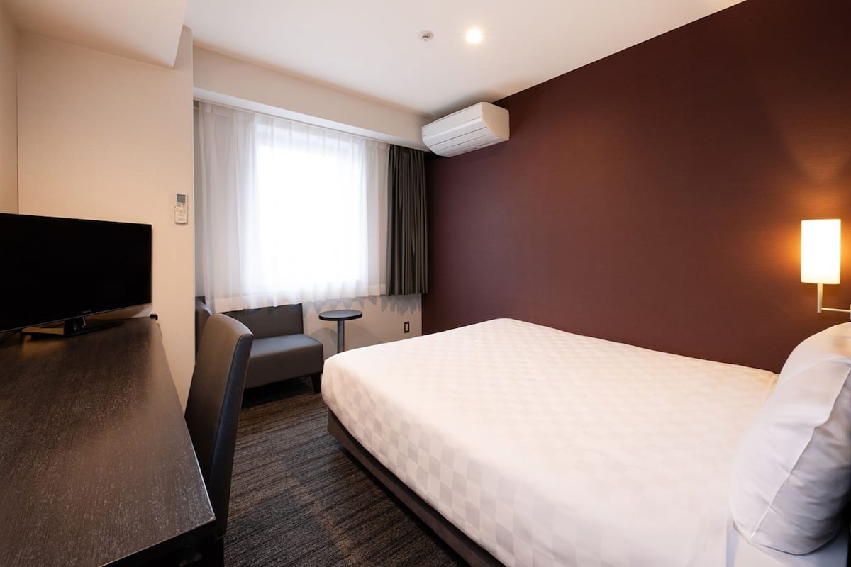 Quarto de hotel com cama, escrivaninha, cadeira, televisão, janela com cortinas, ar condicionado e luminária na parede.