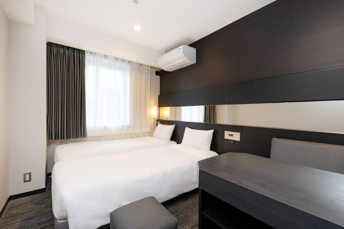Quarto de hotel com duas camas, uma escrivaninha e uma janela grande com cortinas.