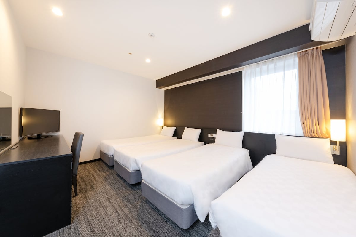 Quarto de hotel com três camas de solteiro, uma escrivaninha com cadeira, televisão de tela plana e uma janela grande com cortinas.