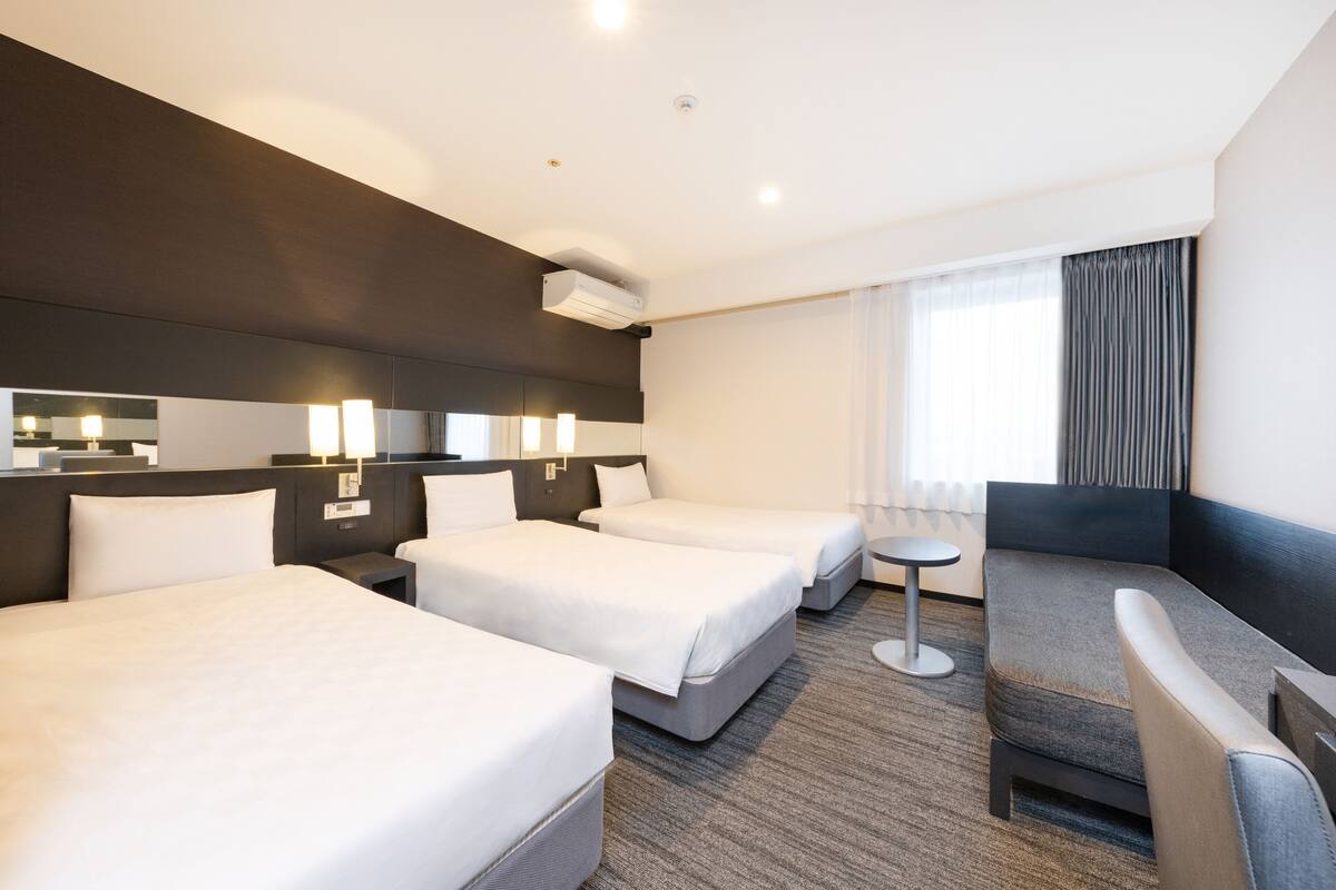 Quarto de hotel com duas camas, um sofá, uma mesinha e uma janela grande com cortinas.