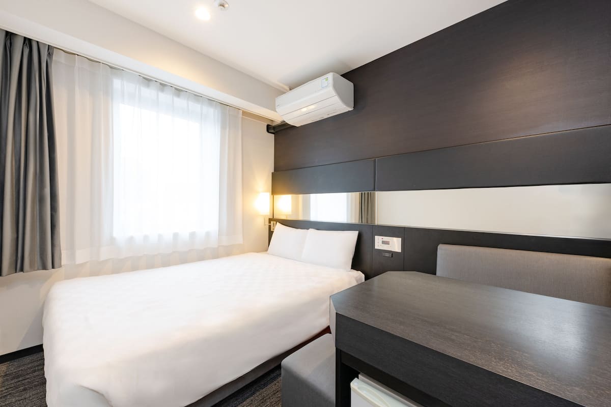 Quarto de hotel com uma cama grande, uma mesa, uma cadeira e ar-condicionado fixado na parede.