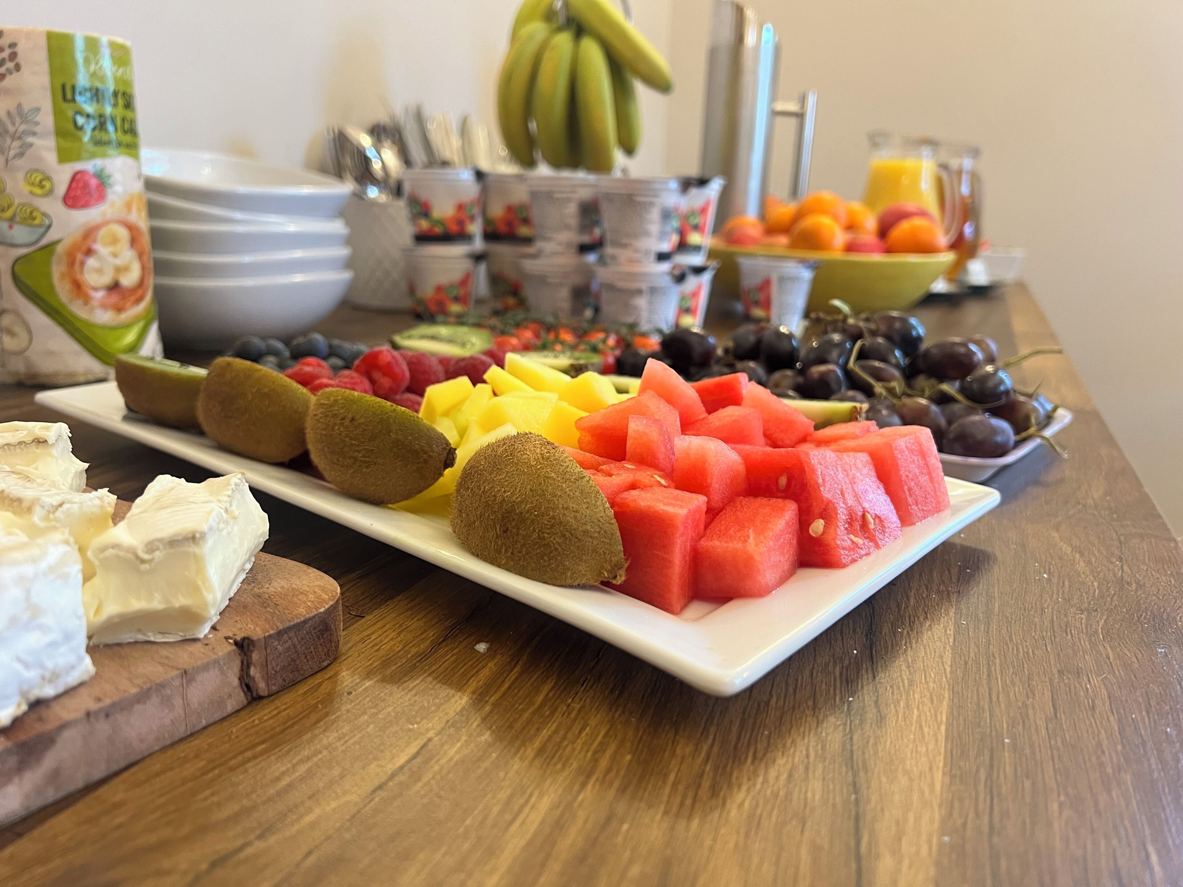 Buffet de café da manhã com diversas frutas, iogurte e queijo sobre uma mesa de madeira.