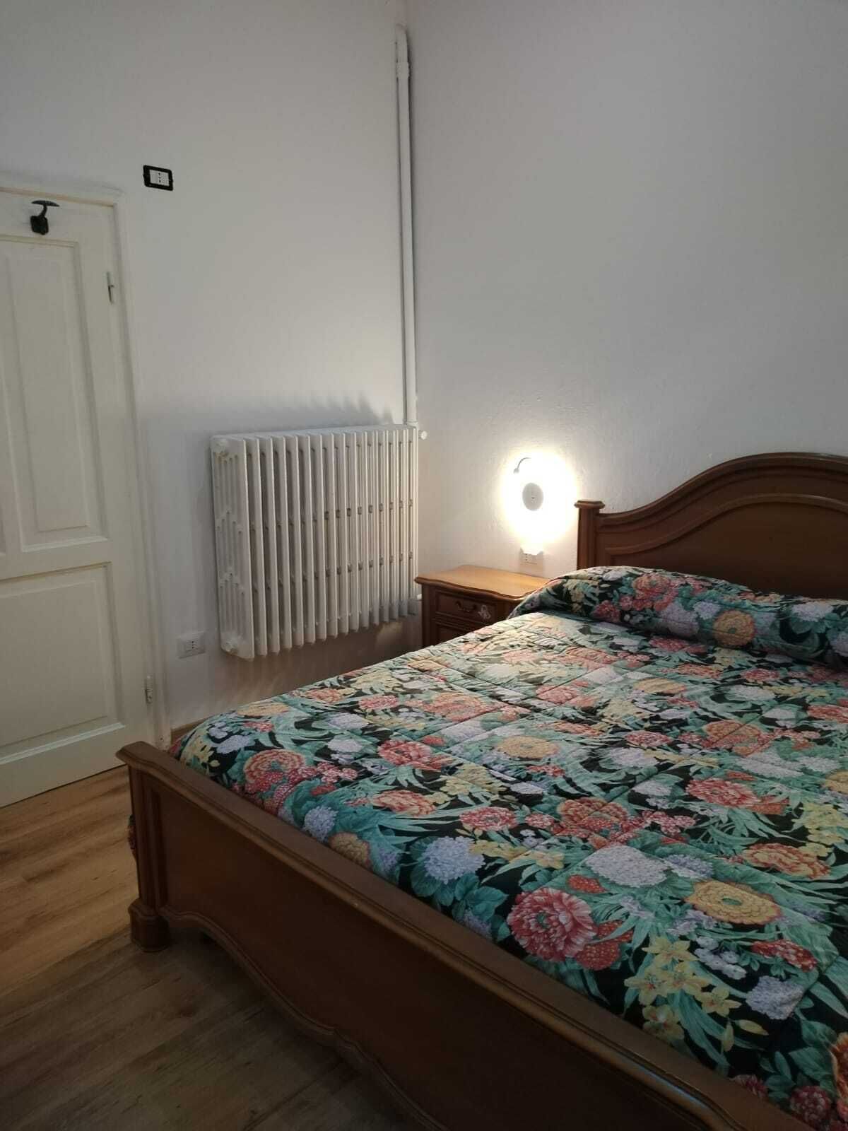 Quarto com cama de madeira, radiador, mesa de cabeceira com abajur e uma porta.