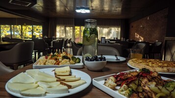 Mesa de jantar com diversos pratos, incluindo pizza, queijo e legumes, em um restaurante com cadeiras e parede de tijolos.