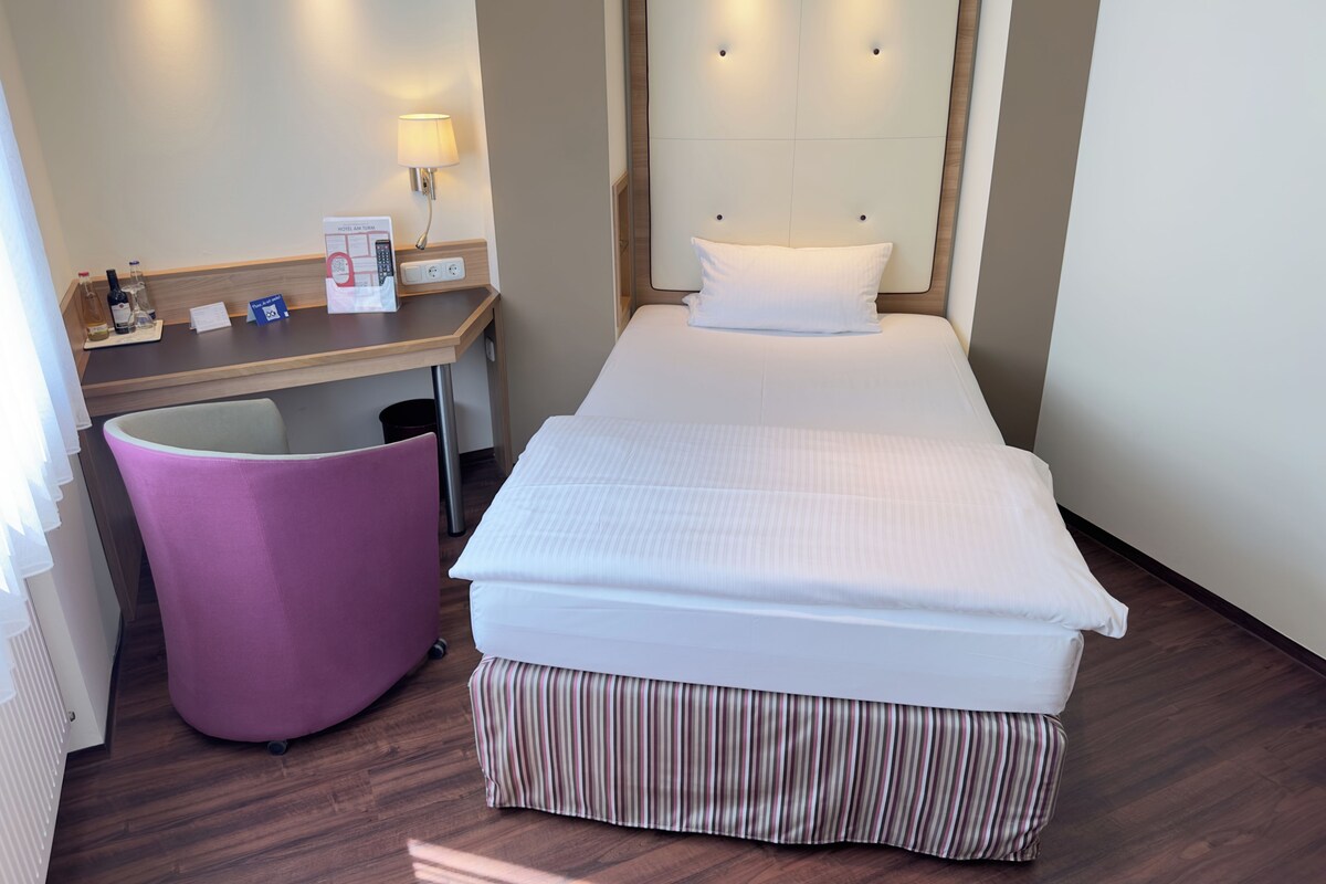 Quarto de hotel com cama, escrivaninha, cadeira, luminária e lixeira.