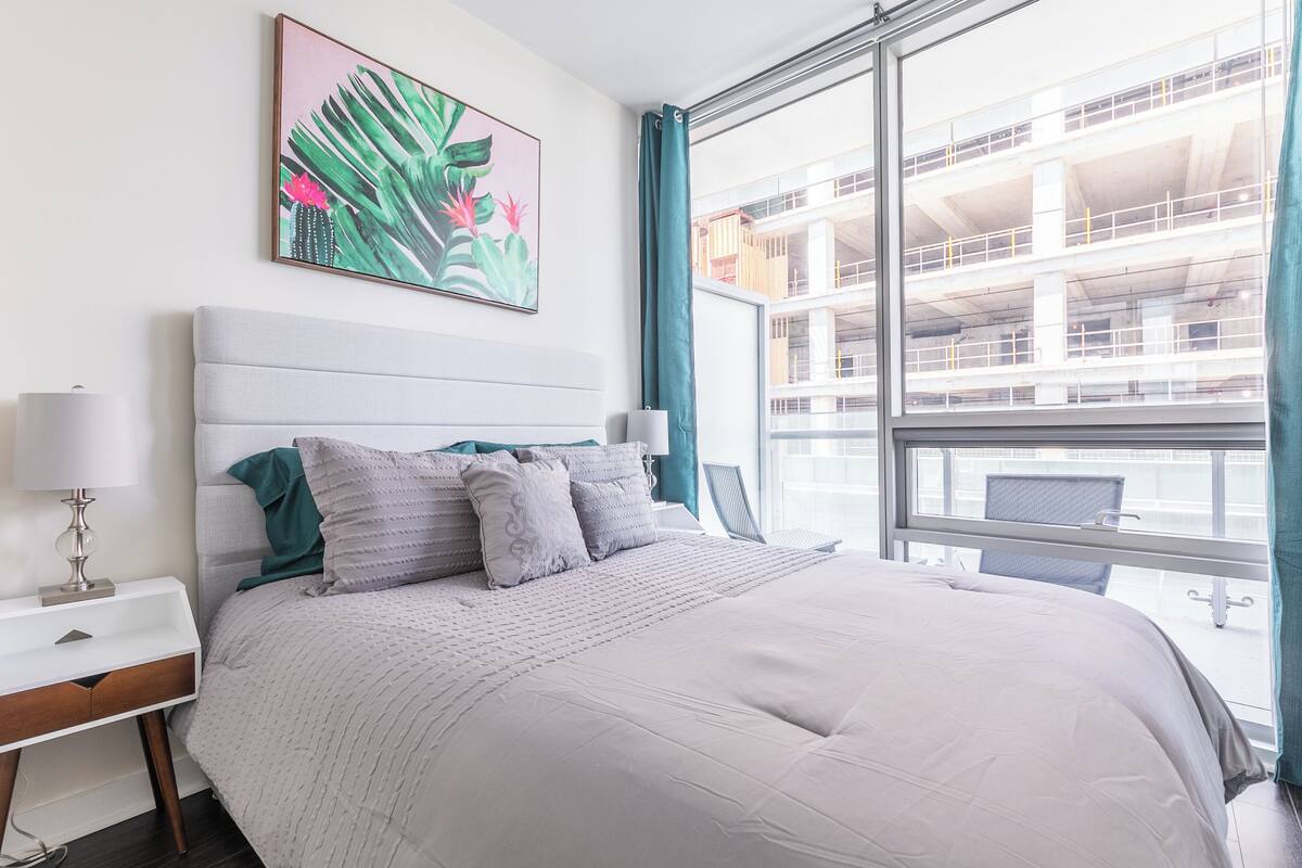 Cama bem arrumada com cabeceira, mesa de cabeceira com abajur e janela com vista para um edifício.