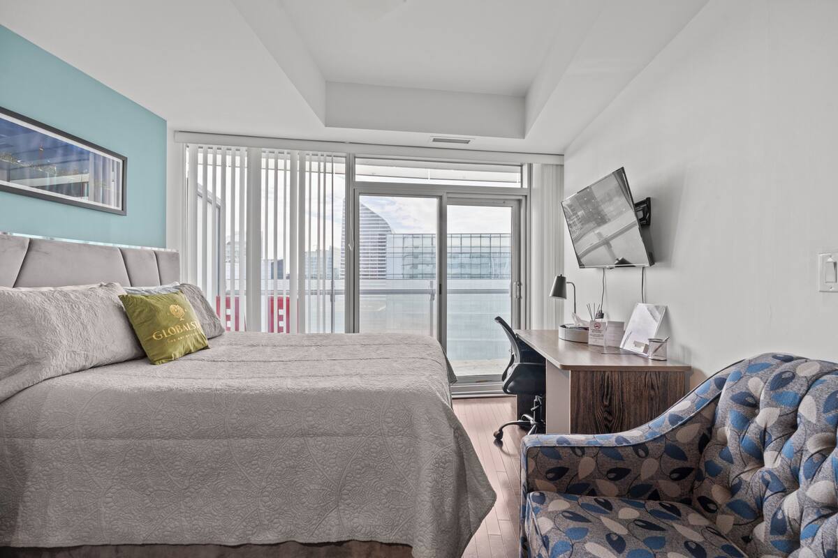 Quarto de hotel com cama, escrivaninha, cadeira, TV e uma grande janela com vista.