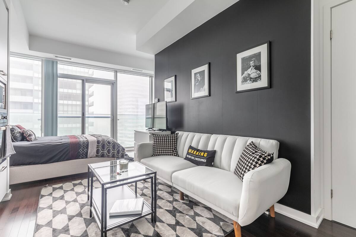 Uma sala de estar moderna com sofá, mesa de centro e cama. Quadros emoldurados decoram a parede e uma grande janela oferece vista da cidade.