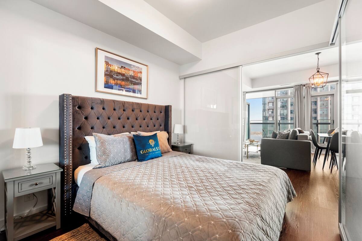 Quarto de hotel moderno com uma cama grande, uma fotografia emoldurada da paisagem urbana, uma pequena mesa com um abajur e vista para a cidade através de grandes janelas.