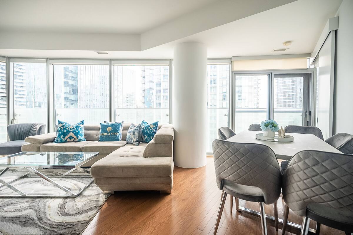 Sala de estar moderna com sofá, mesa de centro e mesa de jantar com cadeiras. Grandes janelas oferecem vista para a cidade.