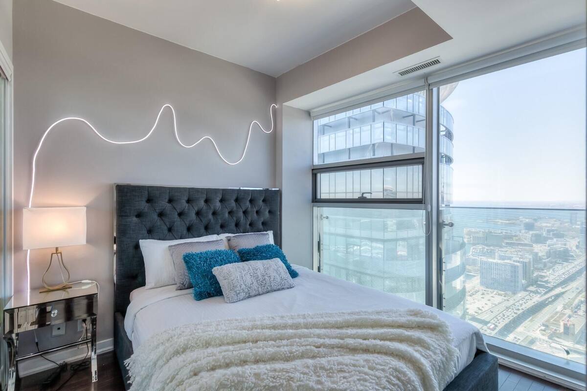 Quarto moderno com uma cama grande, uma mesa de cabeceira, um abajur e vista para a cidade através de uma janela ampla.