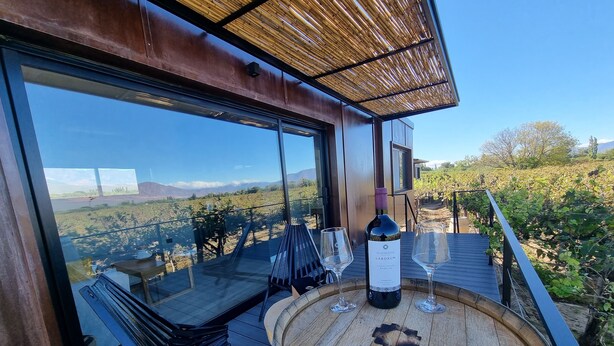 Uma área de estar moderna ao ar livre, com deck de madeira, grandes janelas com vista para um vinhedo e um barril de vinho como peça central.