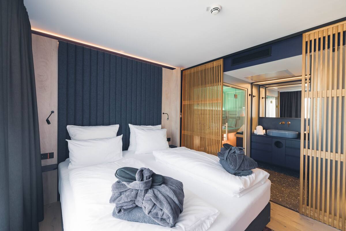 Quarto de hotel moderno com uma cama grande, um guarda-roupa de madeira, um banheiro com pia e espelho, e vista para a cidade.