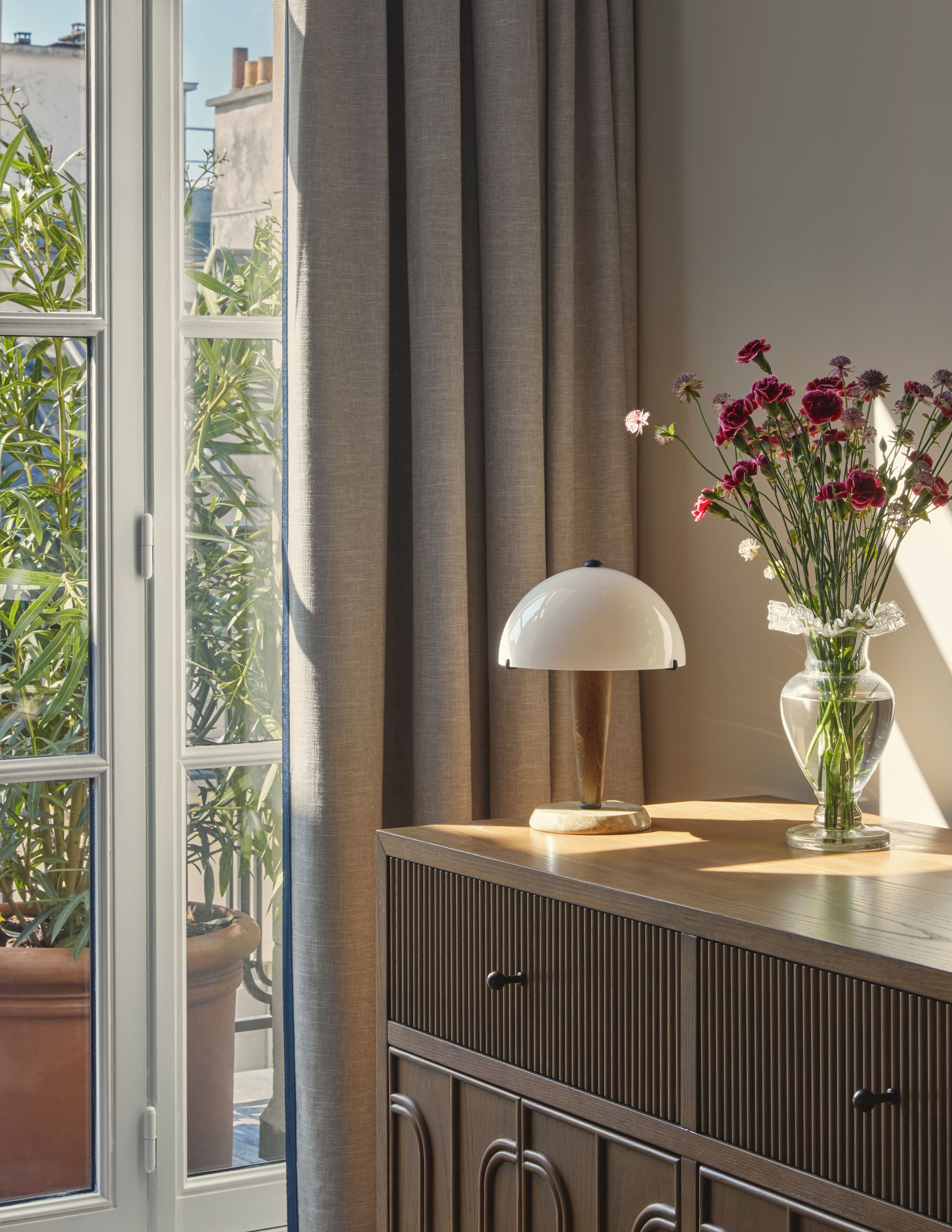 Quarto com vista para vegetação pela janela, abajur sobre mesa de madeira e vaso com flores.
