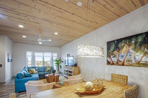 Uma sala de estar ampla com teto de madeira, uma grande mesa de jantar e uma pintura de uma paisagem tropical.