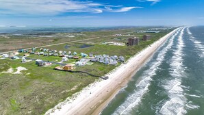 Vista aérea de uma área costeira com uma praia extensa, um pequeno povoado com casas e alguns edifícios.