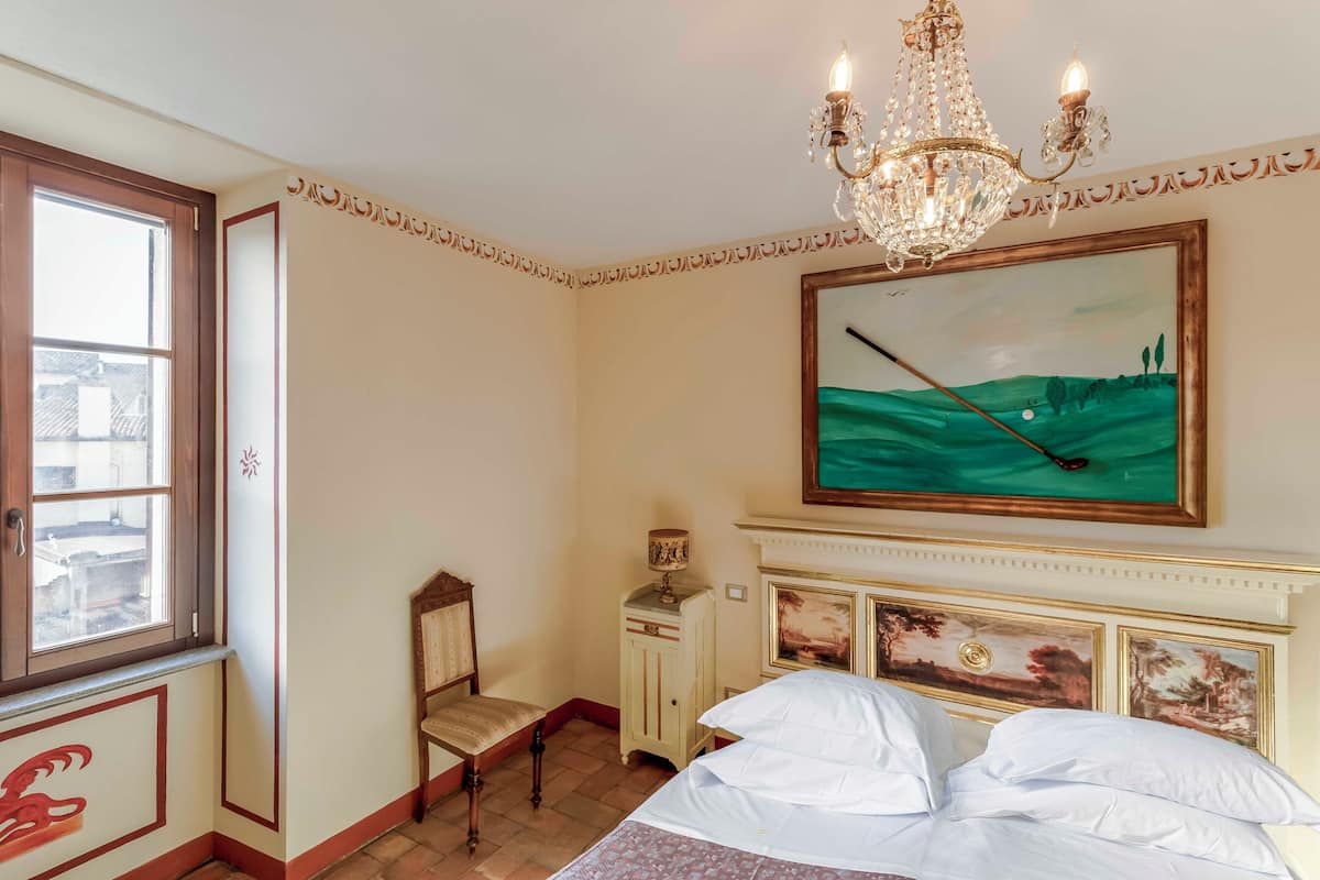 Quarto de hotel com cama, cadeira, armário pequeno, pintura e lustre.