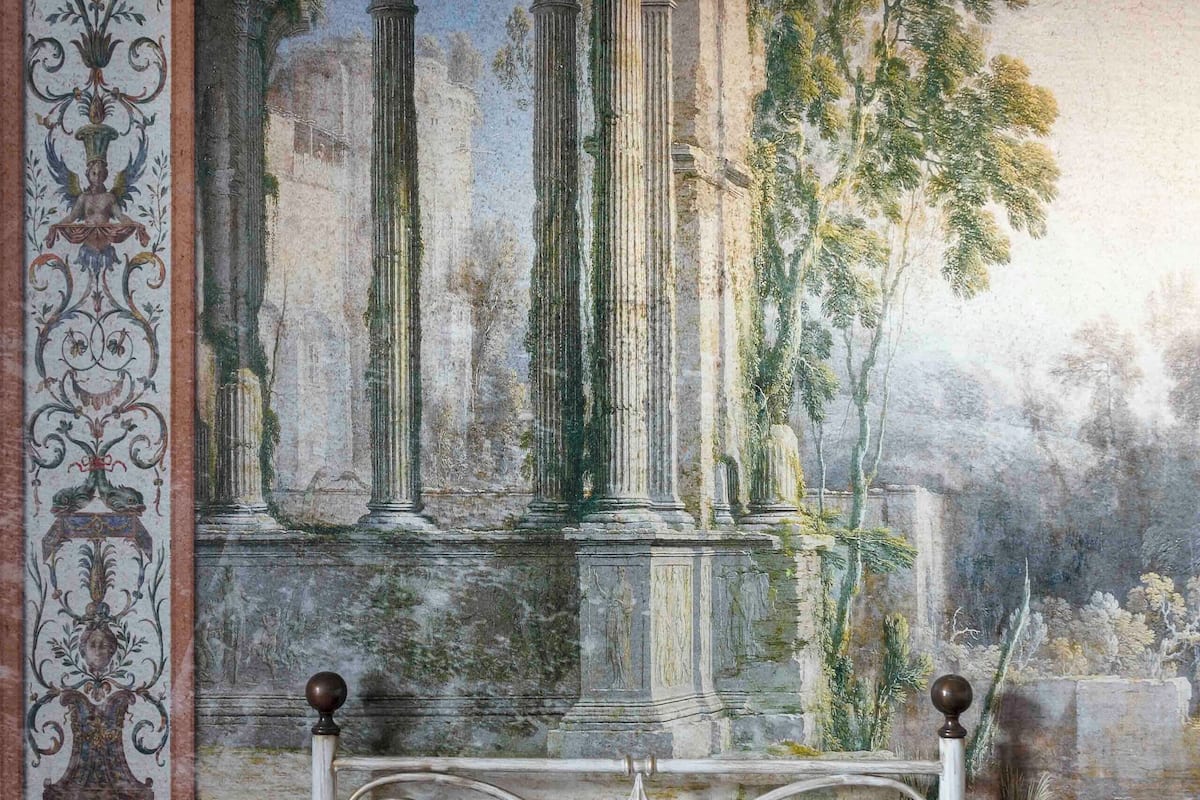 Quarto com uma cama, um mural de colunas antigas e uma moldura decorativa na parede.