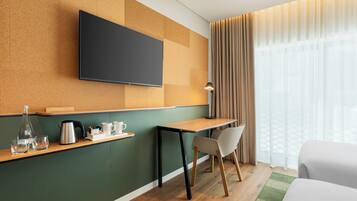 Quarto de hotel moderno com televisão de tela plana presa em parede texturizada, escrivaninha de madeira com cadeira, cafeteira e pequena mesa com xícaras e garrafa.