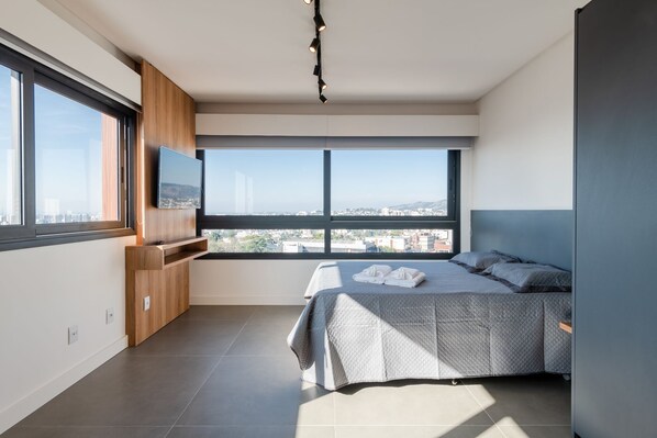 Quarto moderno com uma grande janela, uma cama, uma TV e um armário de madeira.