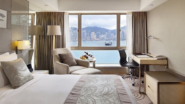 Quarto de hotel com uma cama grande, uma escrivaninha, uma cadeira e vista para a cidade através de grandes janelas.