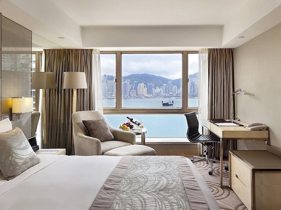Quarto de hotel com uma cama grande, uma escrivaninha, uma cadeira e vista para a cidade através de grandes janelas.