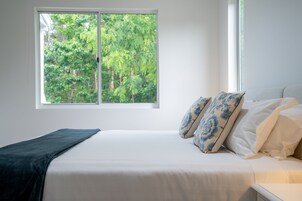Quarto com uma janela grande, cama com roupa de cama branca e almofadas estampadas em azul, e um cobertor escuro no chão.
