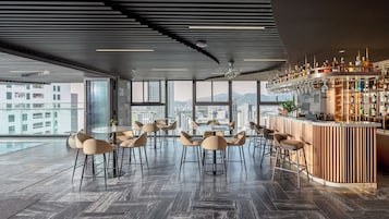 Uma área de bar moderna com vista panorâmica da cidade, apresentando um balcão com diversos tipos de garrafas e copos, e várias mesas redondas com cadeiras.