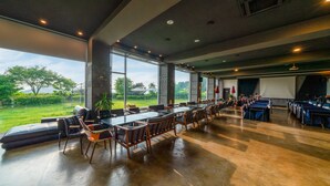 Um espaço amplo e interno com uma mesa comprida e cadeiras, grandes janelas com vista para áreas verdes e um palco com instrumentos musicais.