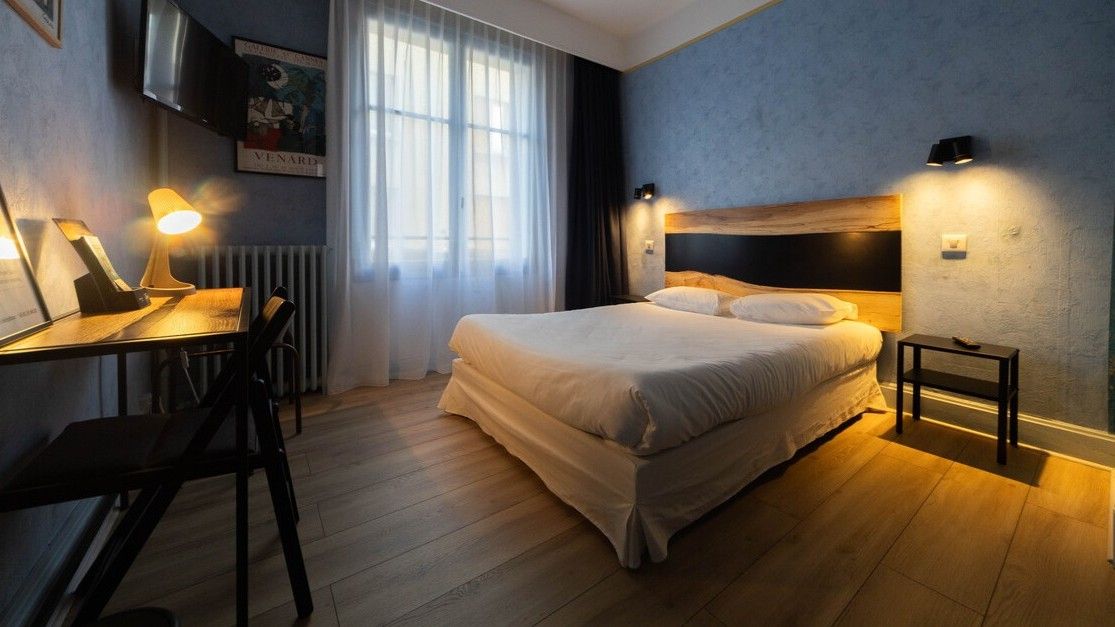 Quarto de hotel com cama, escrivaninha, cadeira, luminária, mesa de cabeceira e janela com cortinas.