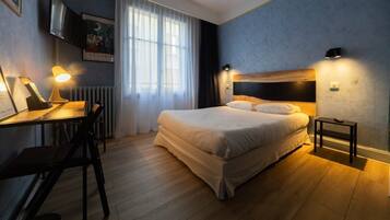 Quarto de hotel com cama, escrivaninha, cadeira, luminária, mesa de cabeceira e janela com cortinas.