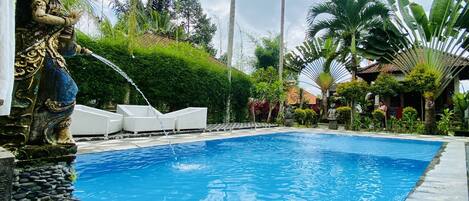 Uma piscina com uma estátua à esquerda, rodeada por palmeiras e um muro de pedra.