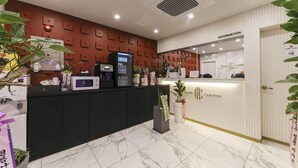 Uma área de recepção de hotel com piso de mármore, balcão de atendimento preto, uma máquina de café e um vaso de plantas.