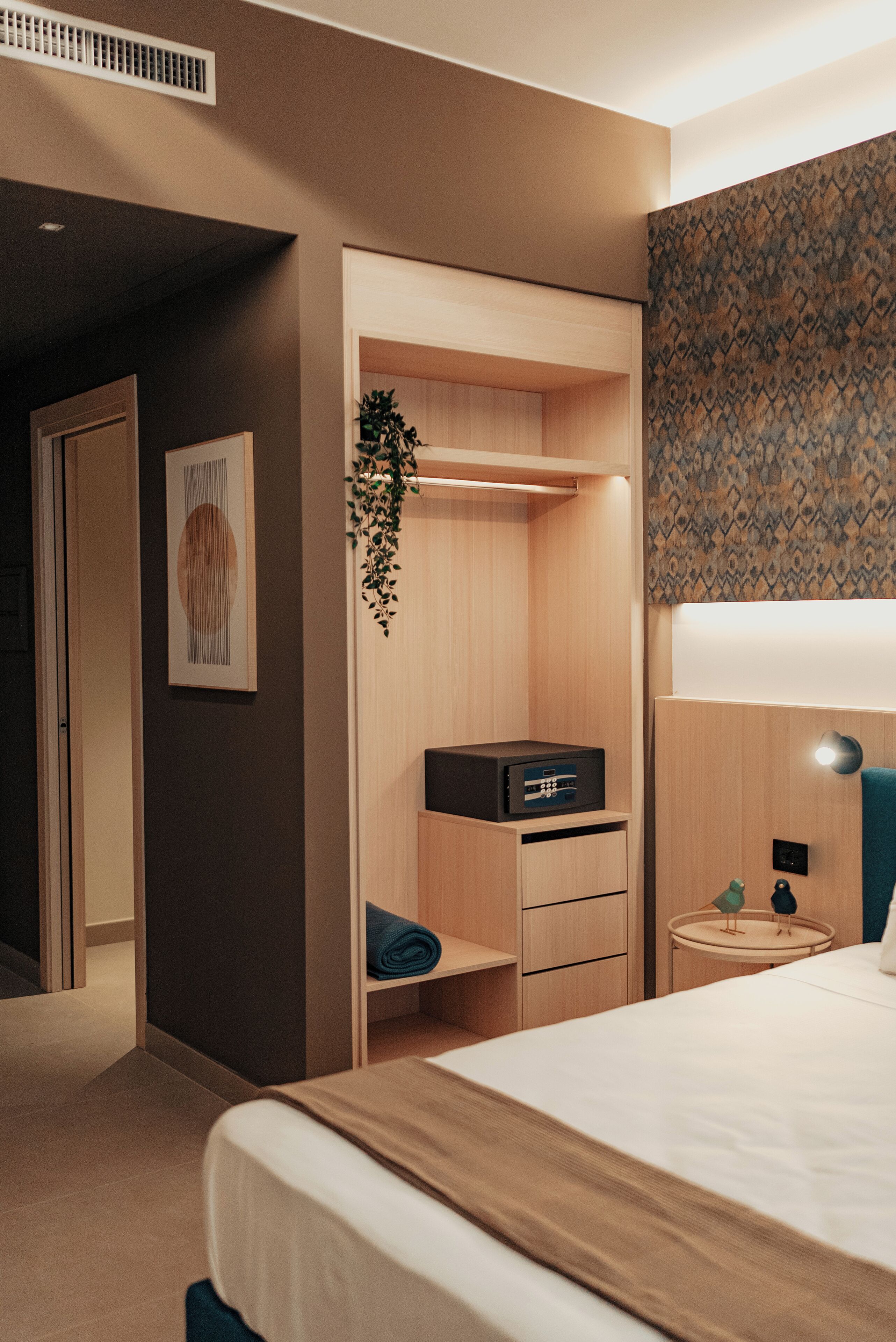 Quarto de hotel com uma cama, um guarda-roupa, uma mesa de cabeceira com luminária e uma pequena planta.