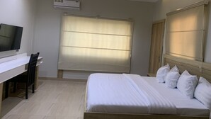 Quarto com cama, escrivaninha, cadeira, televisão e janela com persianas.