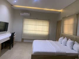 Quarto com cama, escrivaninha, cadeira, televisão e janela com persianas.
