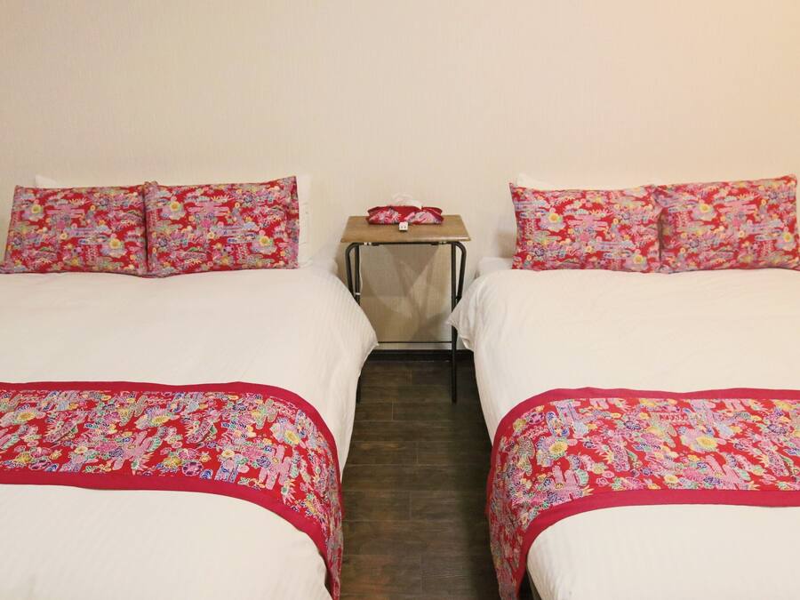 Quarto de hotel com duas camas, cada uma com roupa de cama e almofadas estampadas com padrão floral. Há uma pequena mesa com um tecido vermelho e um objeto branco sobre ela.