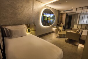 Quarto de hotel moderno com uma cama grande, um espelho redondo, área de estar com mesa de centro e vista para a paisagem urbana pela janela.