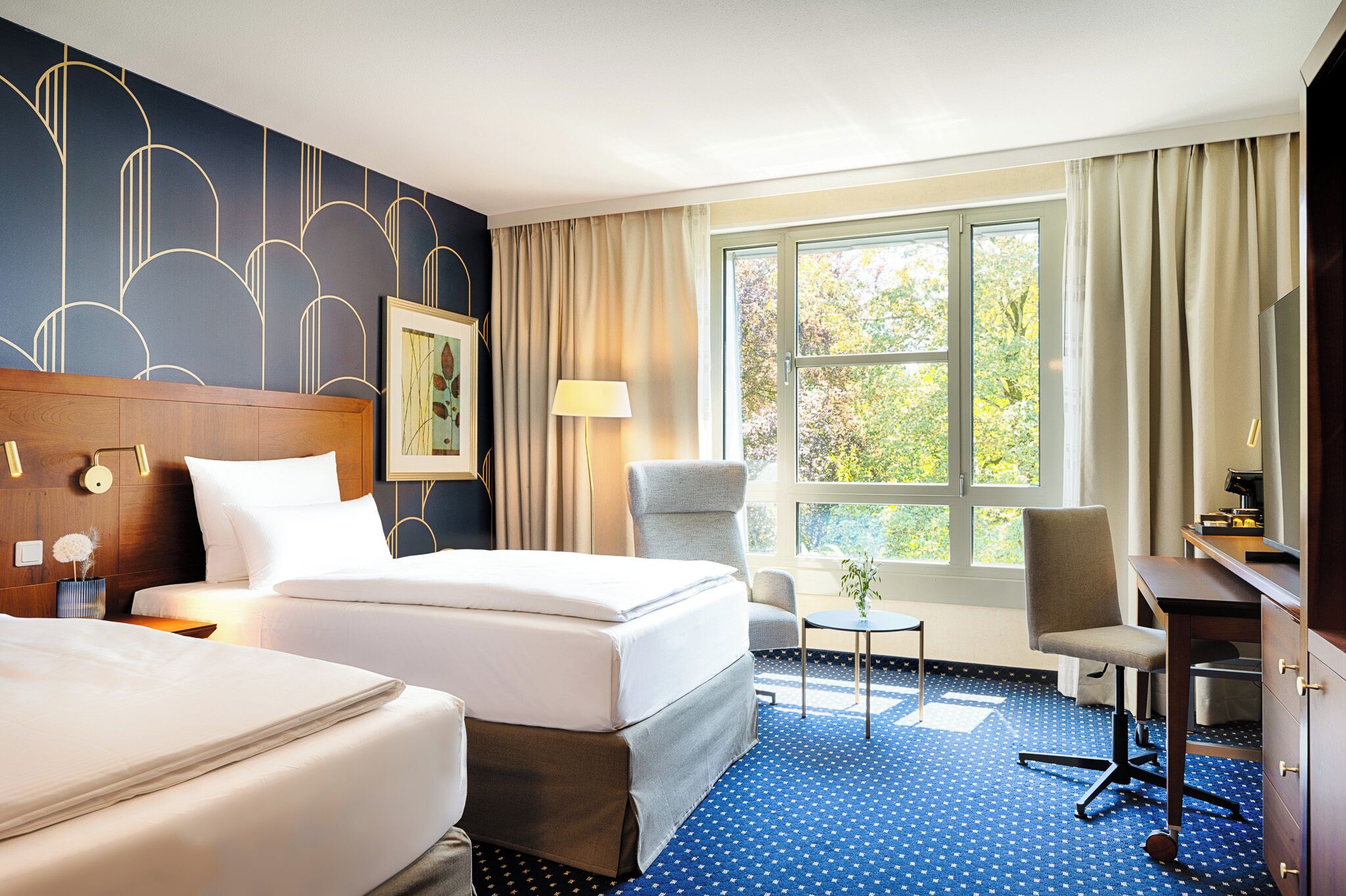 Quarto de hotel com duas camas, uma escrivaninha, uma cadeira, um abajur e uma janela grande com cortinas.