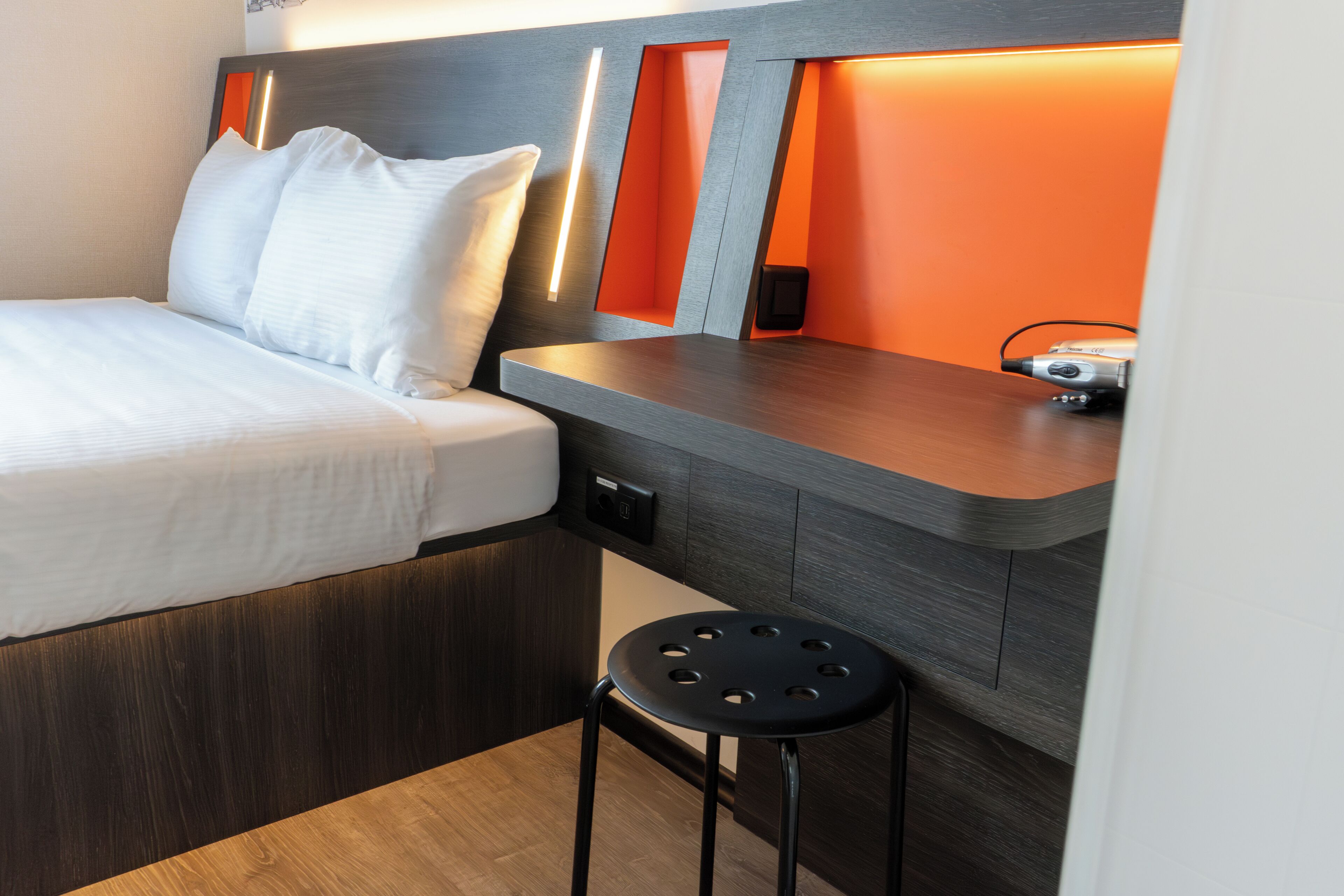 Quarto de hotel moderno com uma cama, um banquinho pequeno e uma escrivaninha com abajur.