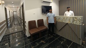 Uma área de recepção de hotel com balcão de mármore, duas cadeiras marrons, uma televisão de tela plana e um longo corredor com portas.