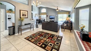 Uma sala de estar espaçosa com um sofá grande, uma mesa de centro e um tapete estampado. Há uma área de jantar com mesa e cadeiras, e uma cozinha com geladeira e lixeira.