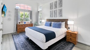 Quarto com cama, mesas de cabeceira, cômoda, janela com cortinas e quadros nas paredes.