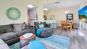 Sala de estar com sofá cinza, mesa de centro e tapete azul. Há também uma área de jantar com mesa e cadeiras de madeira.