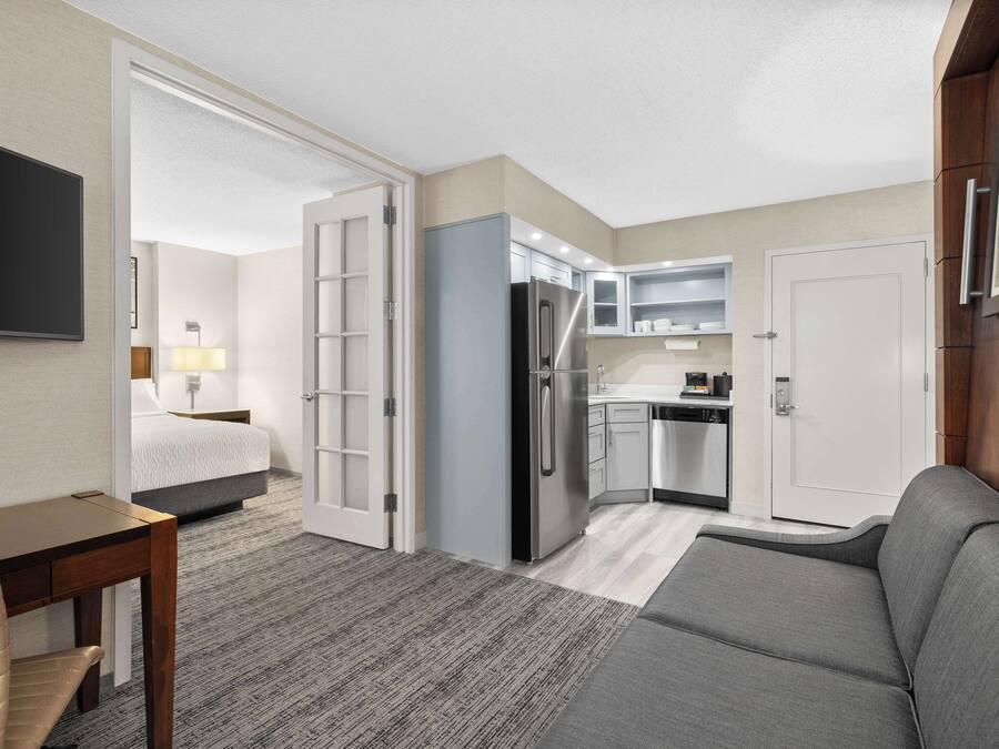 Quarto de hotel com cozinha compacta, sofá cinza, mesa pequena e televisão.