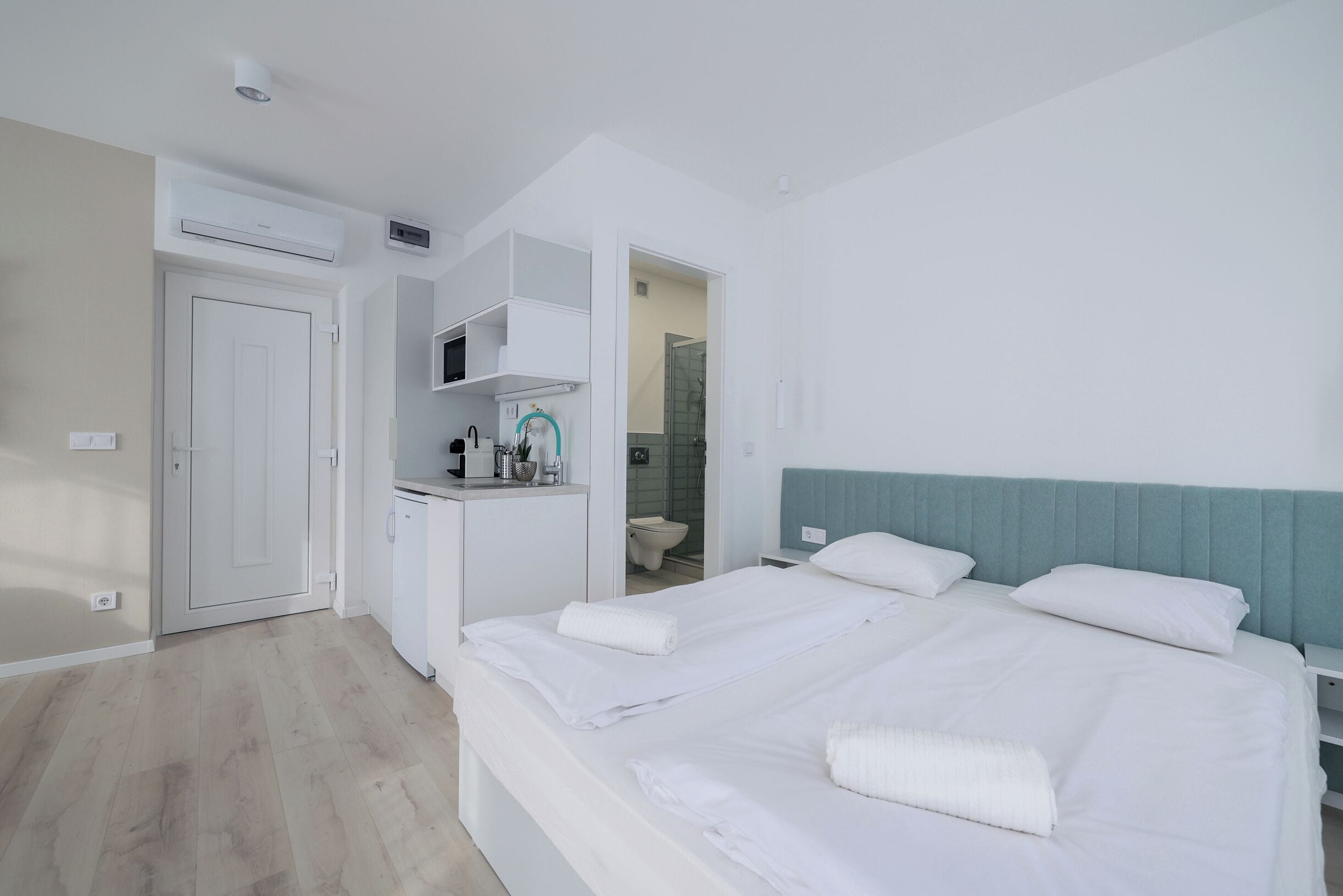 Quarto de hotel moderno com uma cama grande, uma kitchenette pequena e um banheiro visível através de uma porta aberta.