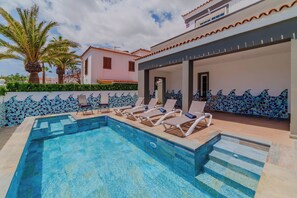 Uma piscina com espreguiçadeiras, um deck revestido de azulejos e um edifício com telhado e paredes azulejadas.