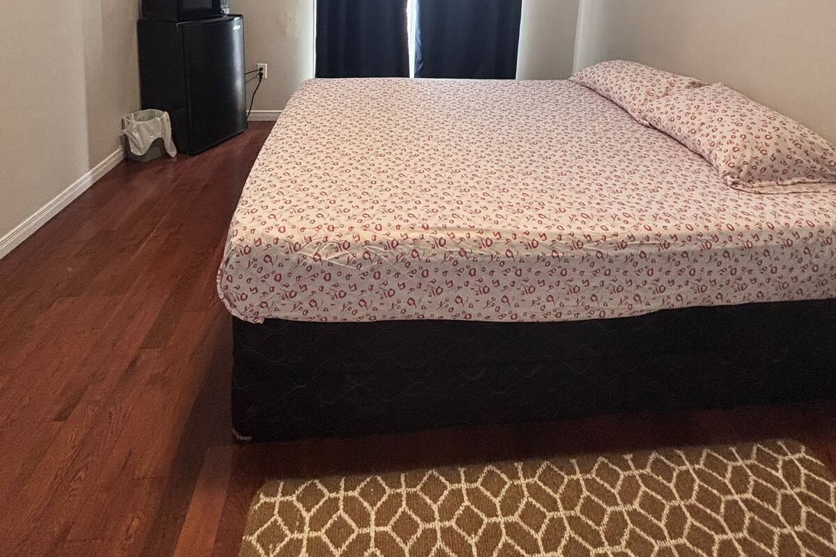 Quarto com cama, uma escrivaninha pequena, uma cadeira, janela com cortinas e luminária no teto.