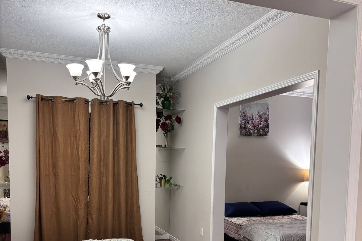 Um quarto com cama, uma cadeira, uma mesinha, uma estante com plantas e um quadro na parede.